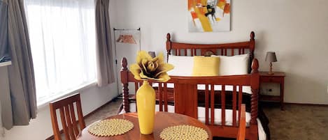 Quarto com uma cama de madeira, uma mesa redonda de madeira com um vaso amarelo e um quadro na parede.