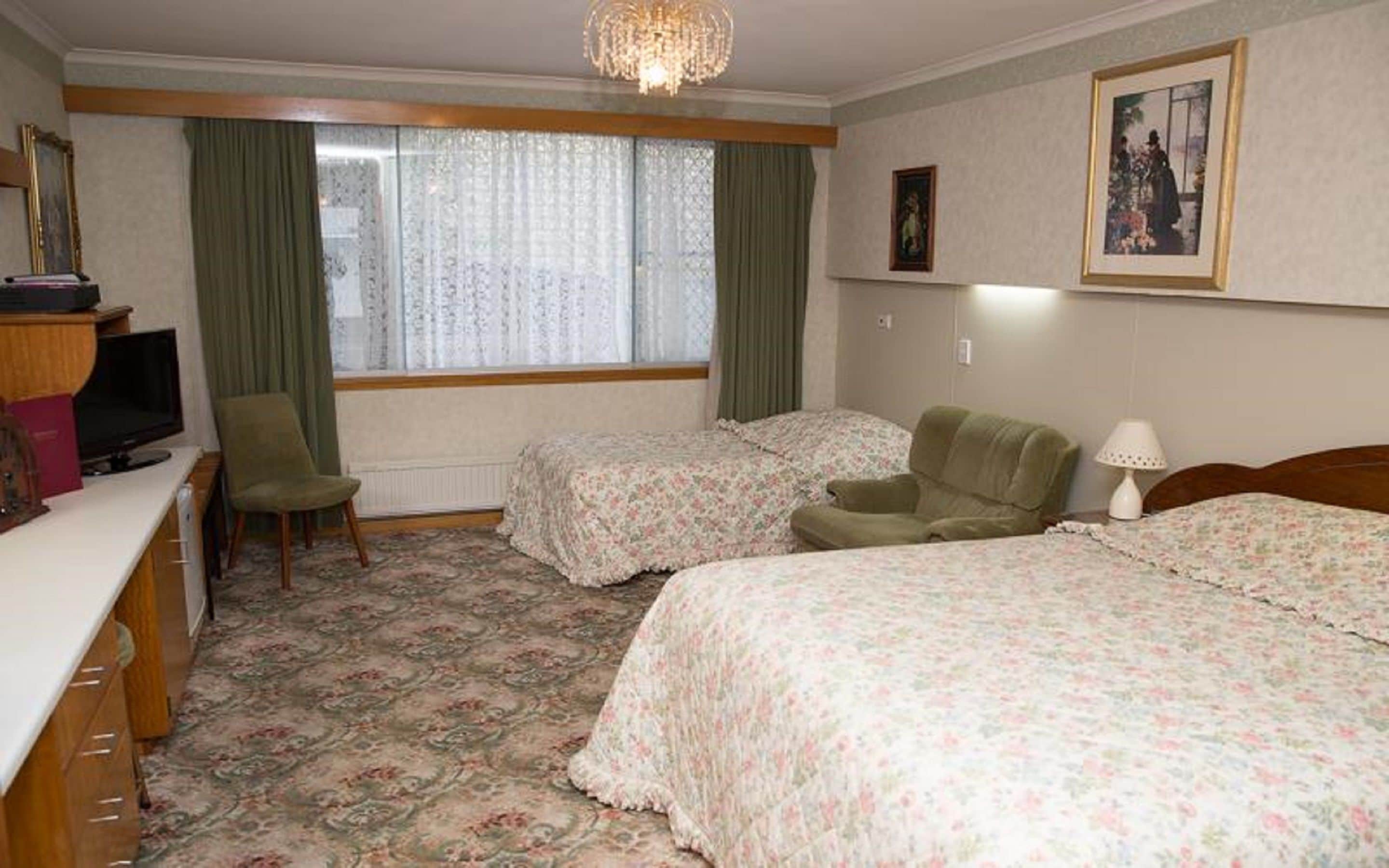 Quarto de hotel com duas camas de solteiro, uma escrivaninha com televisão, uma cadeira e uma janela com cortinas.
