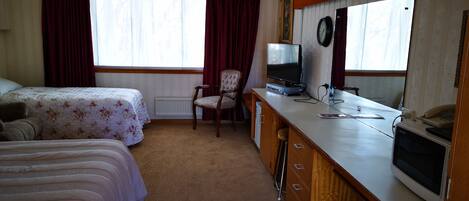 Quarto de hotel com duas camas, uma escrivaninha com televisão, uma cadeira e uma janela grande com cortinas.