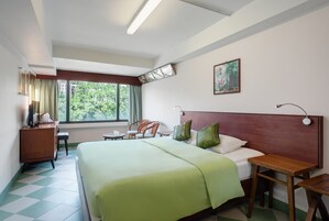 Quarto de hotel com uma cama grande, uma escrivaninha, uma cadeira e uma janela com vista para árvores.