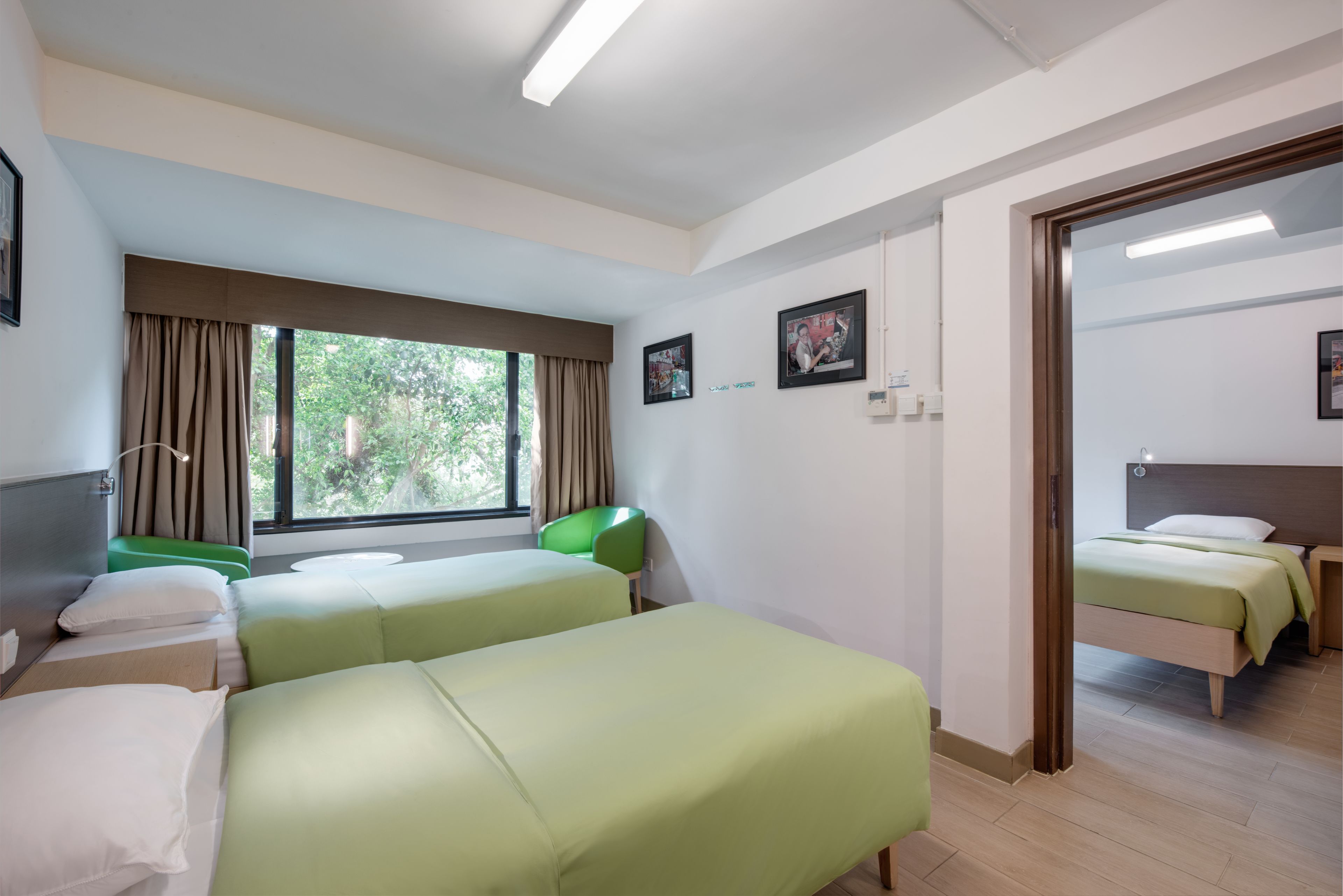 Quarto de hotel com duas camas, uma janela com cortinas, uma cadeira verde e quadros nas paredes.