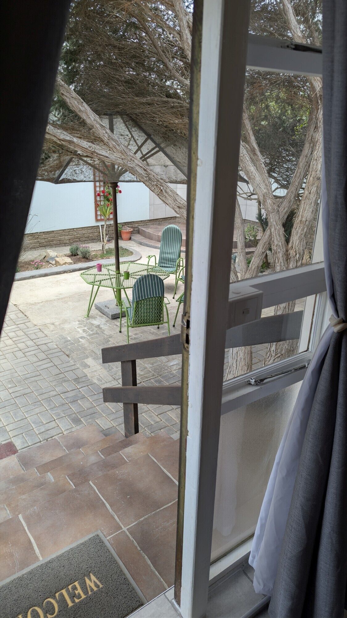 Vista da janela de um quarto, com porta, tapete de boas-vindas e um espaço de pátio com cadeiras e mesa.