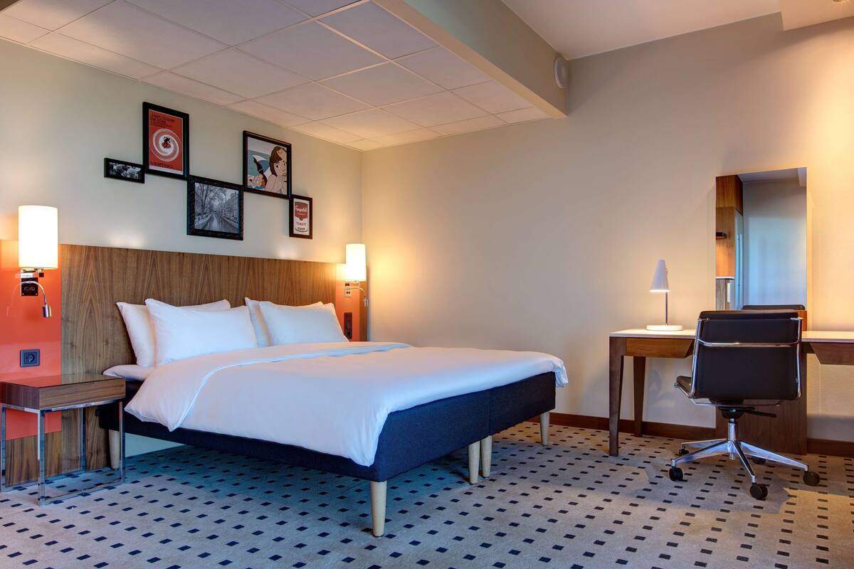 Quarto de hotel com cama, escrivaninha, cadeira e luminárias fixadas na parede.