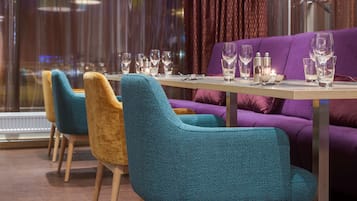 Uma área de refeições moderna com uma mesa posta para dois, um sofá roxo e cadeiras verde-água.