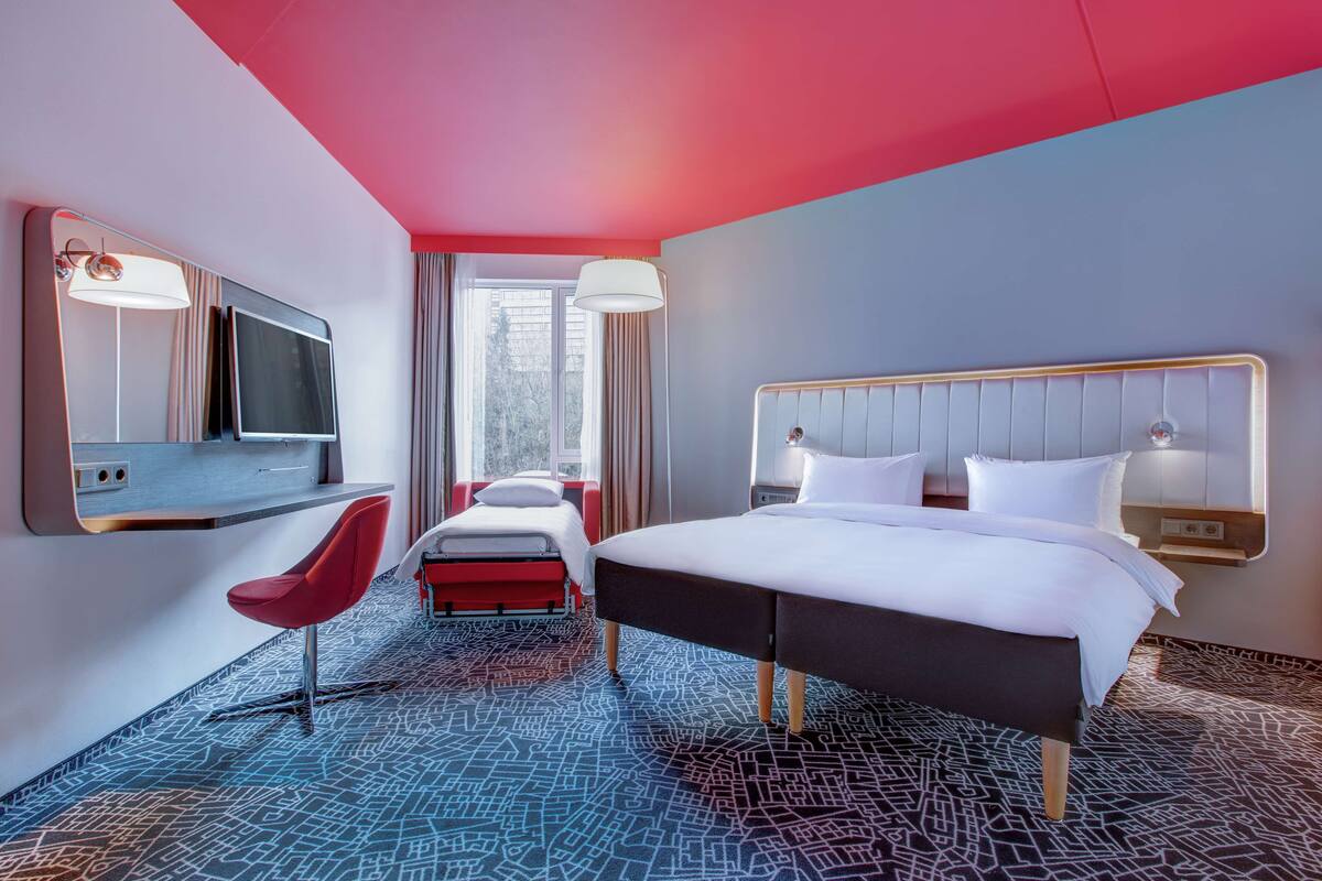 Quarto de hotel com uma cama grande, uma escrivaninha com cadeira, televisão e janela com cortinas.