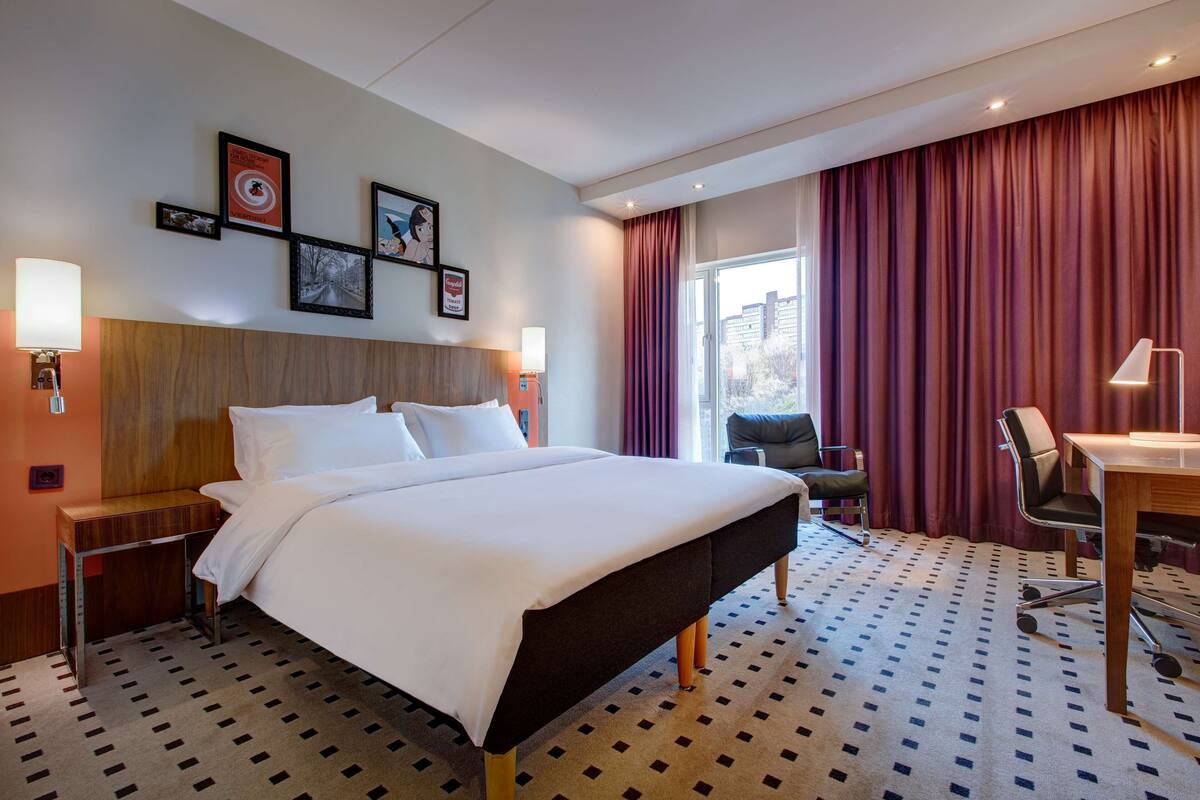 Quarto de hotel com uma cama grande, uma escrivaninha e uma cadeira. Há luminárias na parede e uma janela com cortinas.