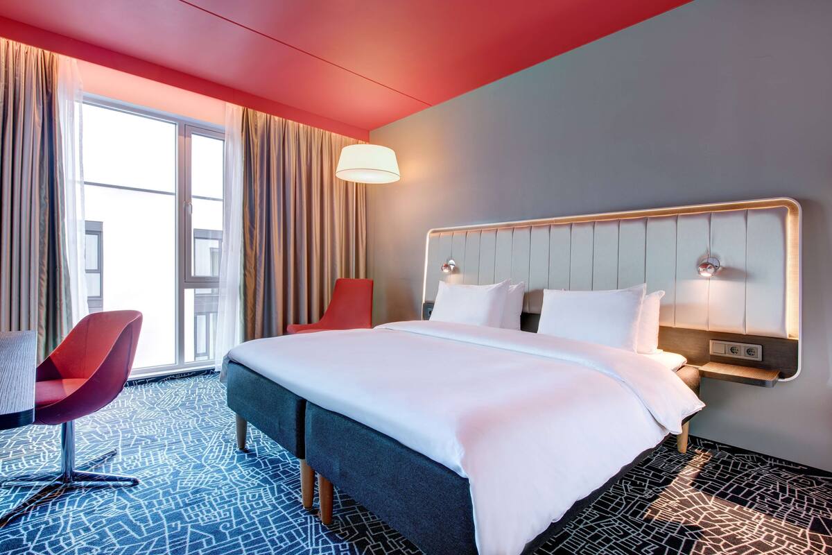 Quarto de hotel com uma cama grande, uma cadeira vermelha, uma escrivaninha e uma janela com cortinas.