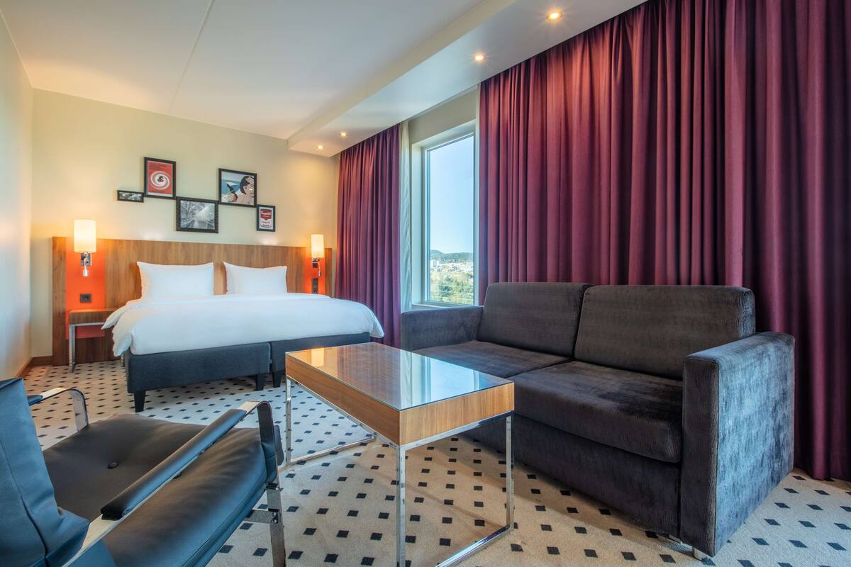 Quarto de hotel com cama, sofá, mesa com tampo de vidro e uma cadeira. Há duas luminárias fixadas na parede e uma janela com cortinas.