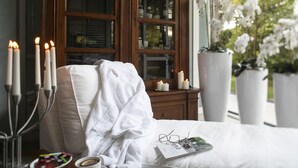 Um quarto aconchegante com uma cama, uma mesa de cabeceira com uma xícara de café, um livro e uma revista. Há velas na mesa e uma janela com vista para vegetação.