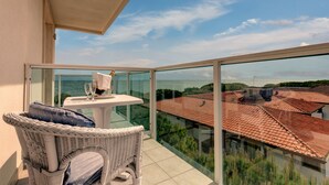 Uma varanda com uma cadeira de vime, uma mesa pequena e uma porta de vidro que dá para um terraço com vista para edifícios e áreas verdes.
