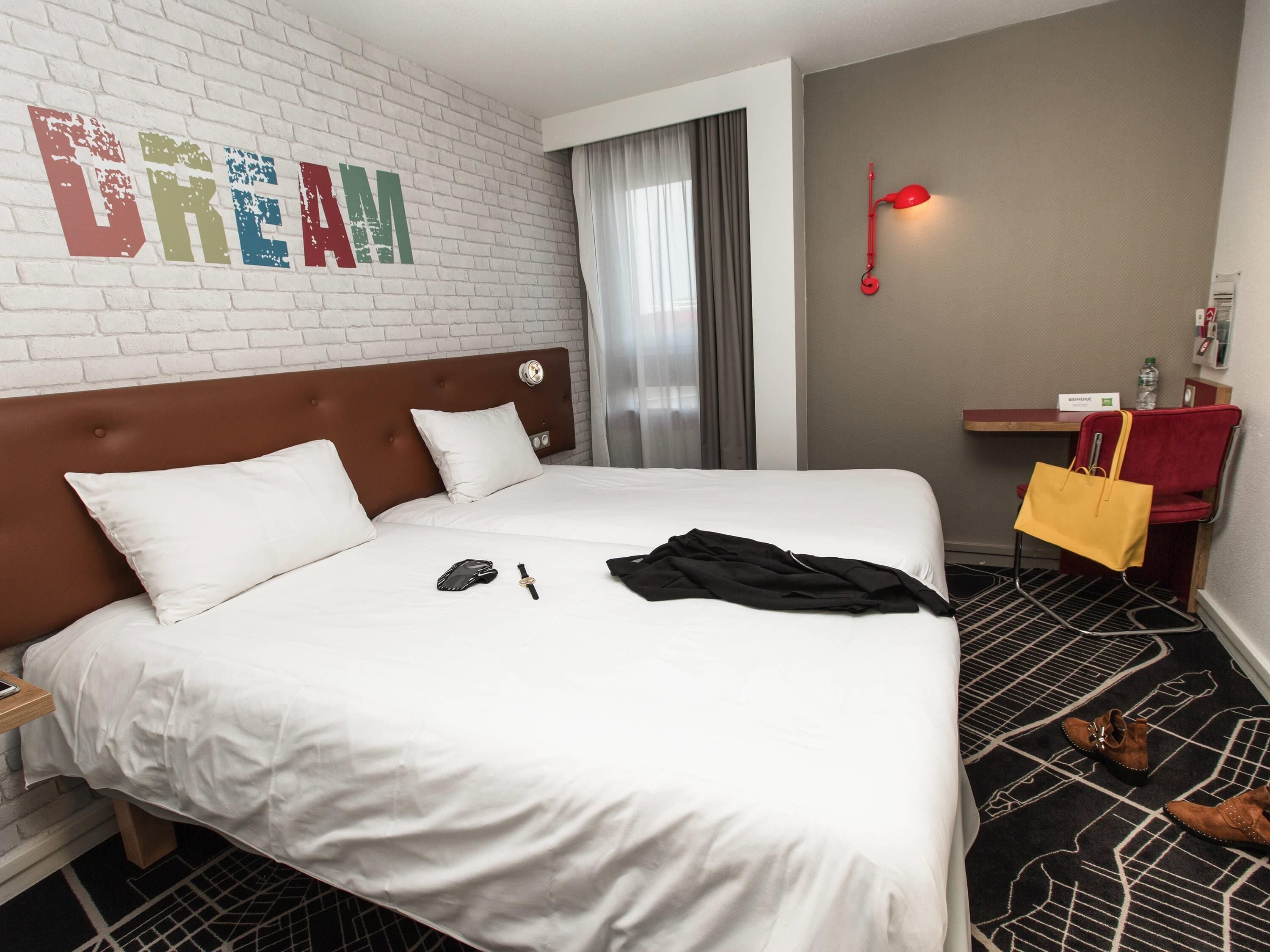 Quarto de hotel com parede de tijolos brancos, cabeceira marrom, edredom branco, luminária vermelha, cadeira vermelha e tapete com padrão preto e branco.
