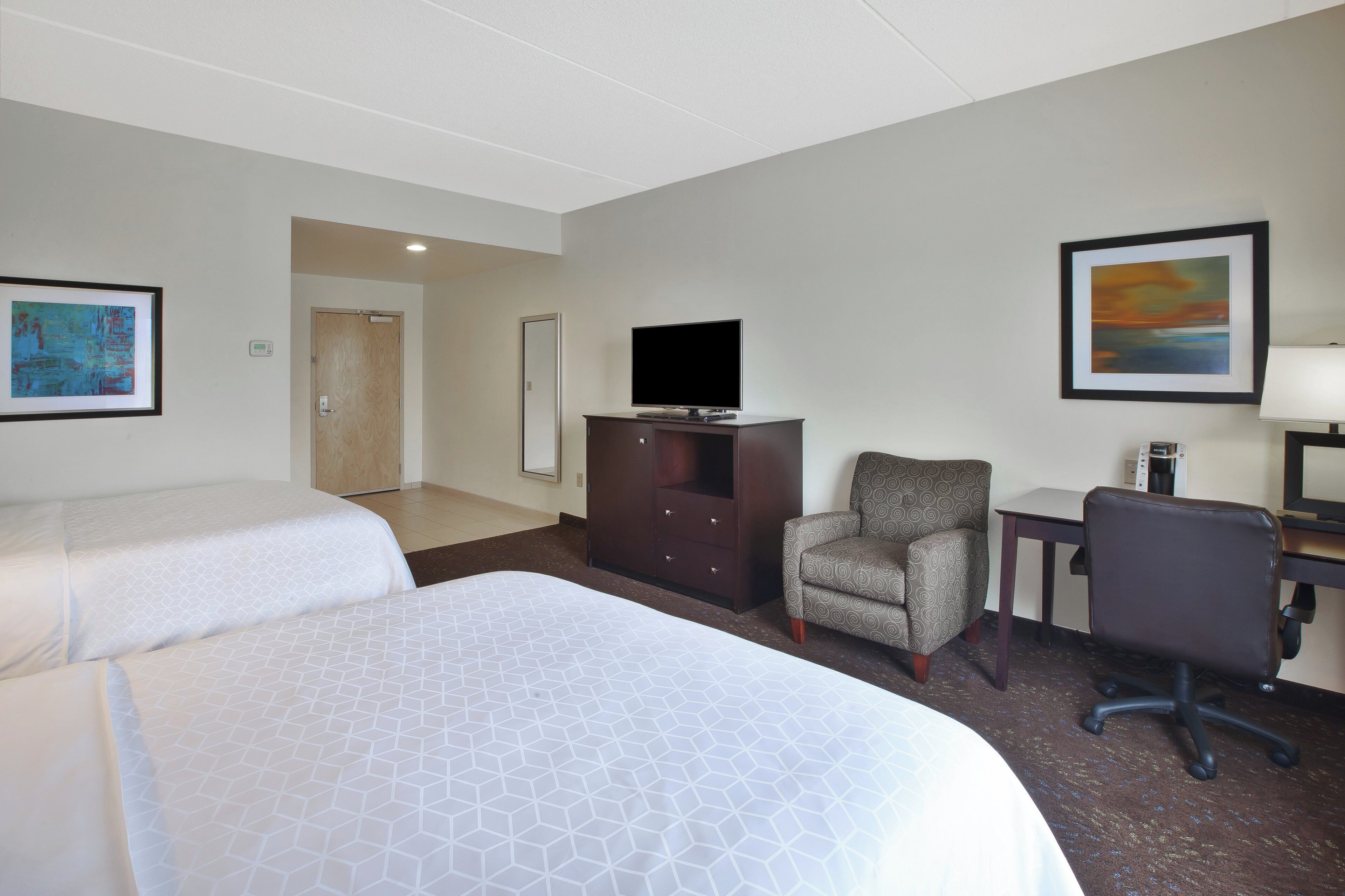 Quarto de hotel com duas camas, uma escrivaninha, uma cadeira, televisão e obras de arte nas paredes.