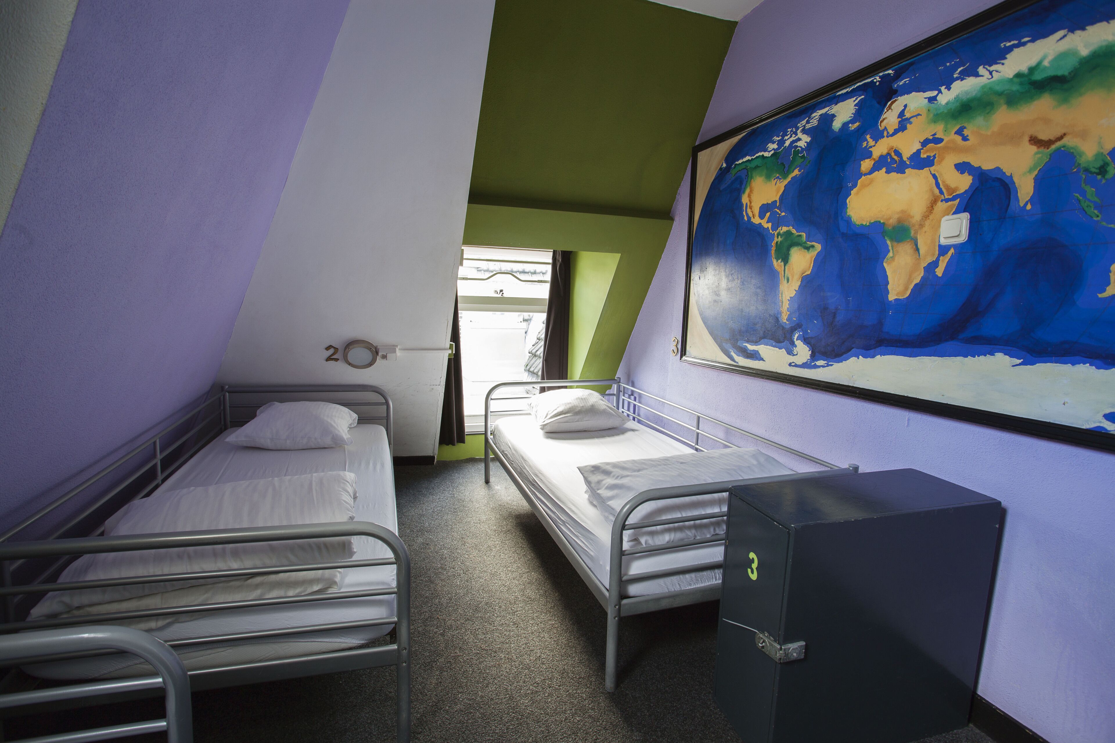 Quarto com duas beliches de metal, uma parede de destaque verde, um grande mapa-múndi e um pequeno armário preto.