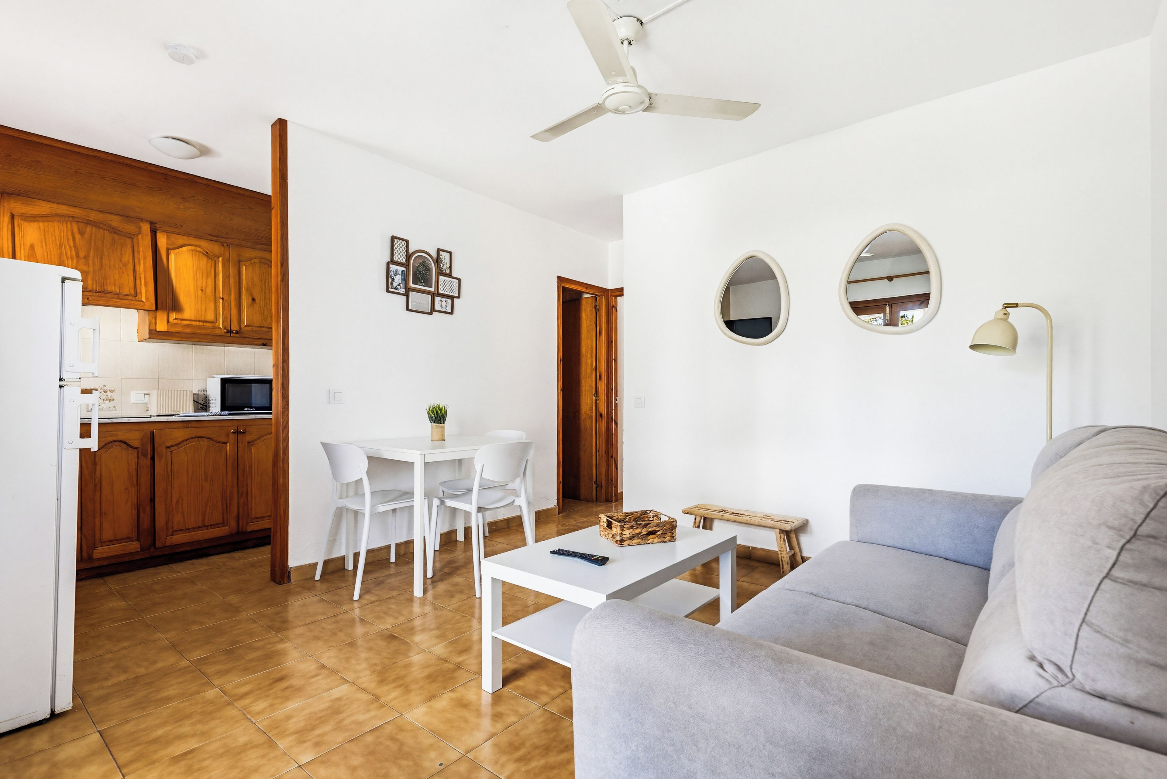 Uma sala de estar moderna com um sofá cinza, uma área de jantar com mesa e cadeiras brancas, uma cozinha com armários de madeira e um ventilador de teto.