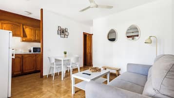 Uma sala de estar moderna com um sofá cinza, uma área de jantar com mesa e cadeiras brancas, uma cozinha com armários de madeira e um ventilador de teto.