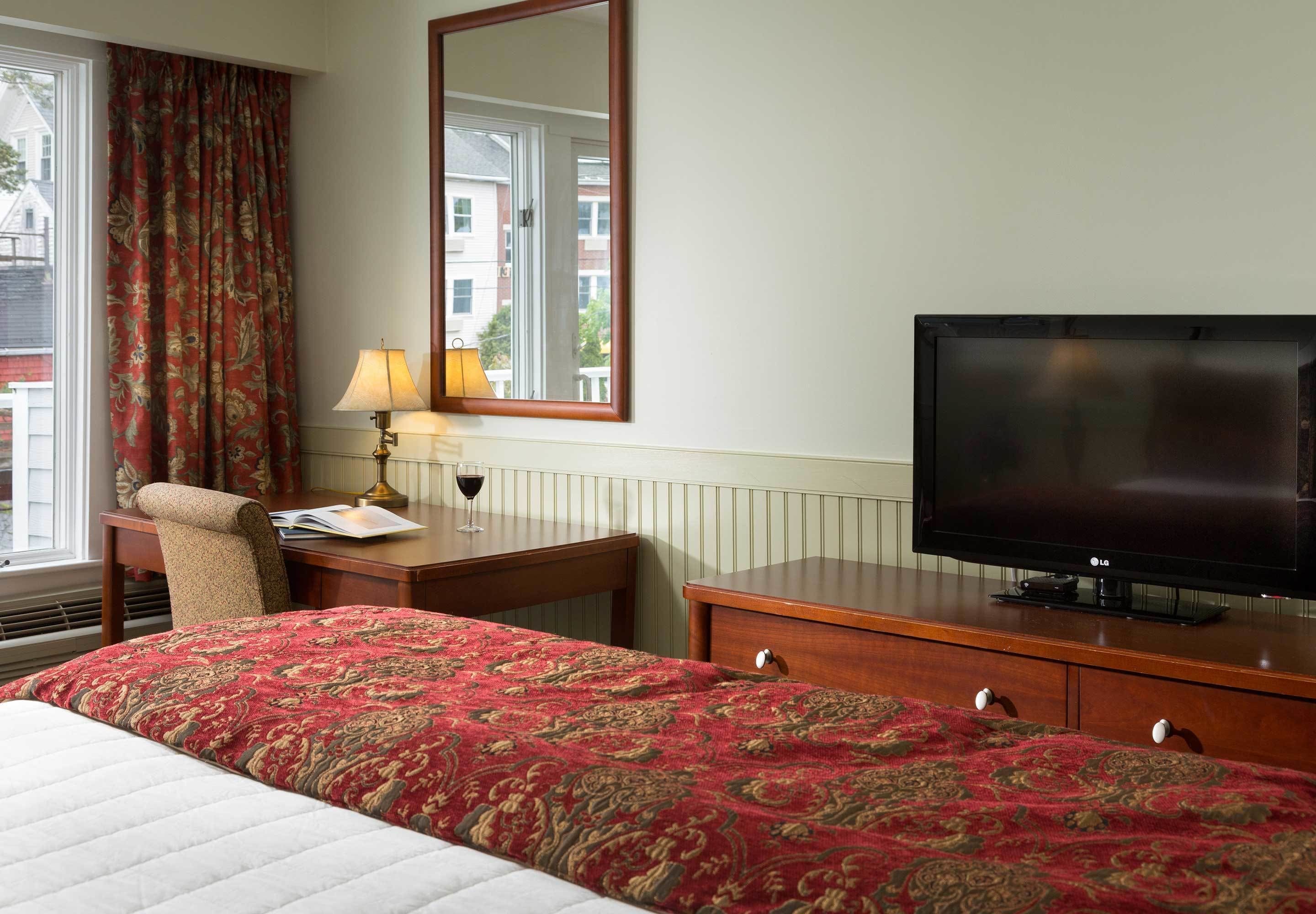 Quarto de hotel com cama, escrivaninha iluminada por uma luminária, televisão e janela com cortinas.