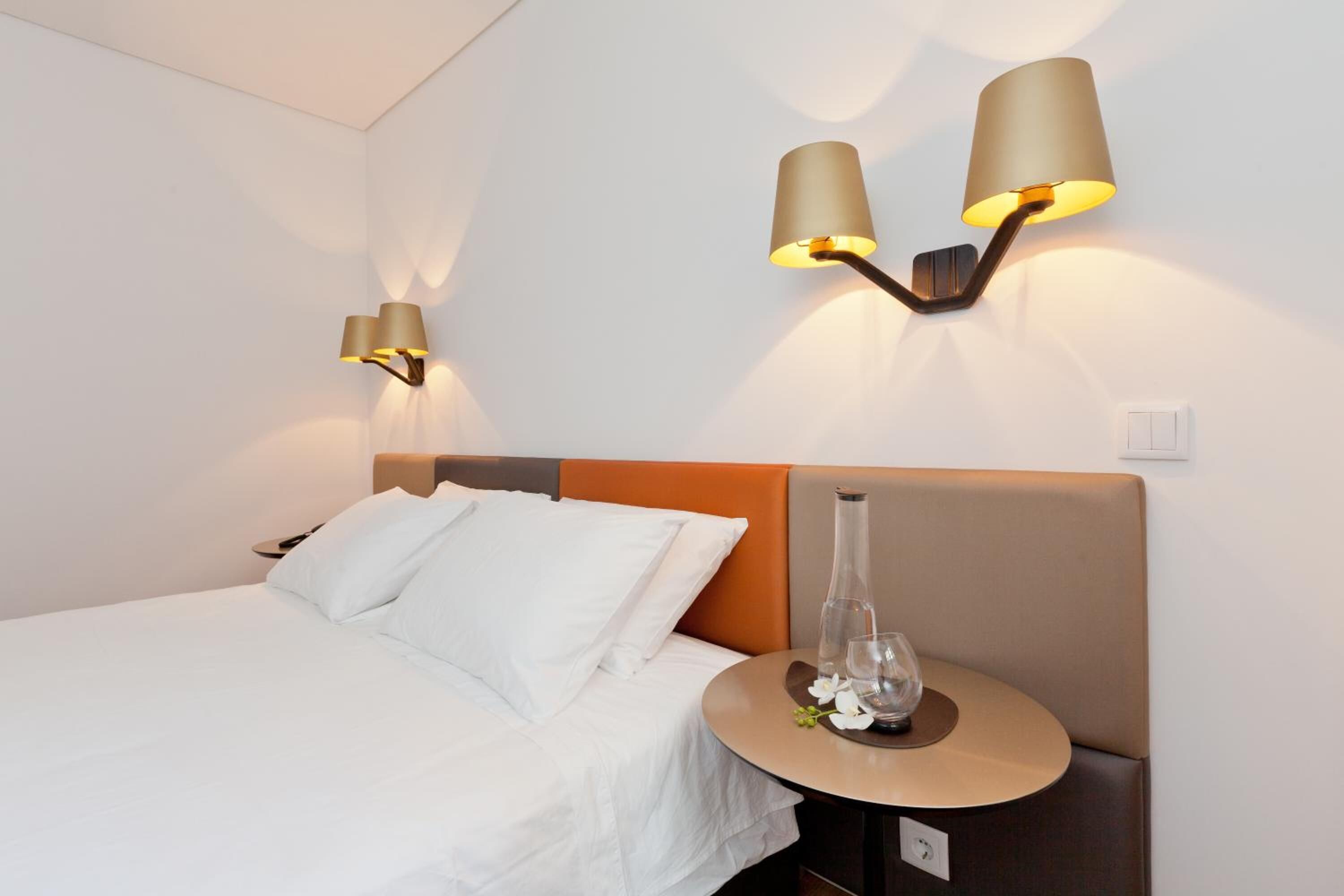 Quarto de hotel com uma cama, duas luminárias fixadas na parede, uma mesa de cabeceira com uma garrafa e copos, e um relógio.