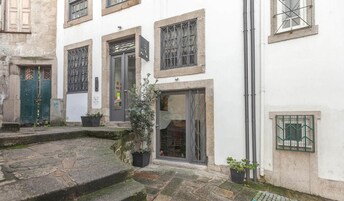 Edifício com fachada de pedra, várias janelas com grades de metal e um pequeno jardim com vasos de plantas.