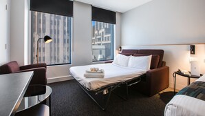 Quarto de hotel com cama, escrivaninha, cadeira e janela com persianas.