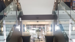 Entrada de hotel com tapete de boas-vindas, portas de vidro e escada.