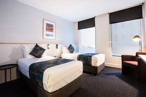 Quarto de hotel com duas camas, uma escrivaninha, uma cadeira, um quadro na parede e uma janela com vista para prédios.