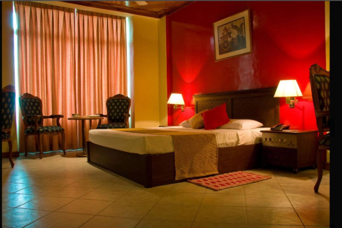 Quarto de hotel com uma cama, duas cadeiras, uma mesinha de cabeceira e uma parede vermelha.