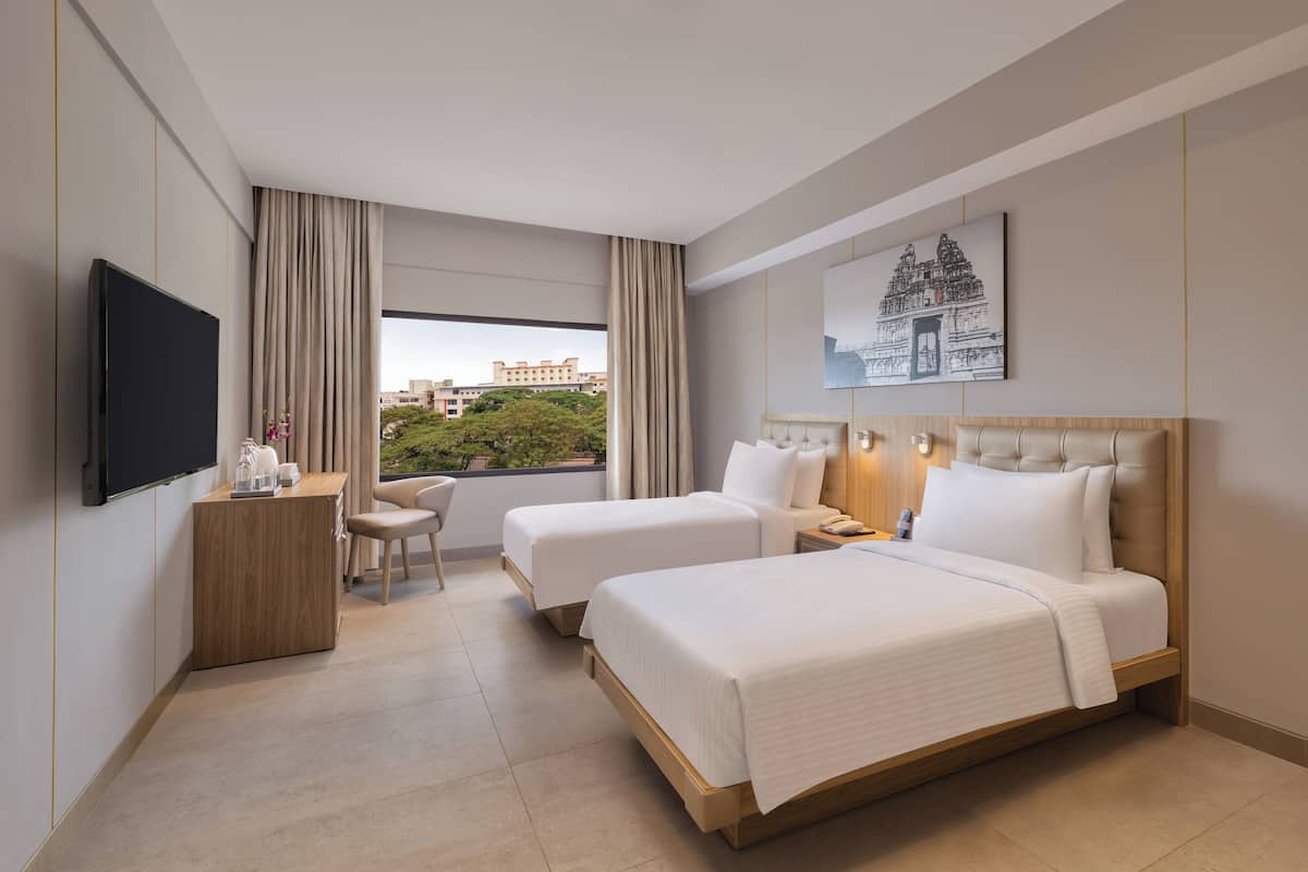 Quarto de hotel com duas camas, televisão, escrivaninha e janela com vista para edifícios e vegetação.