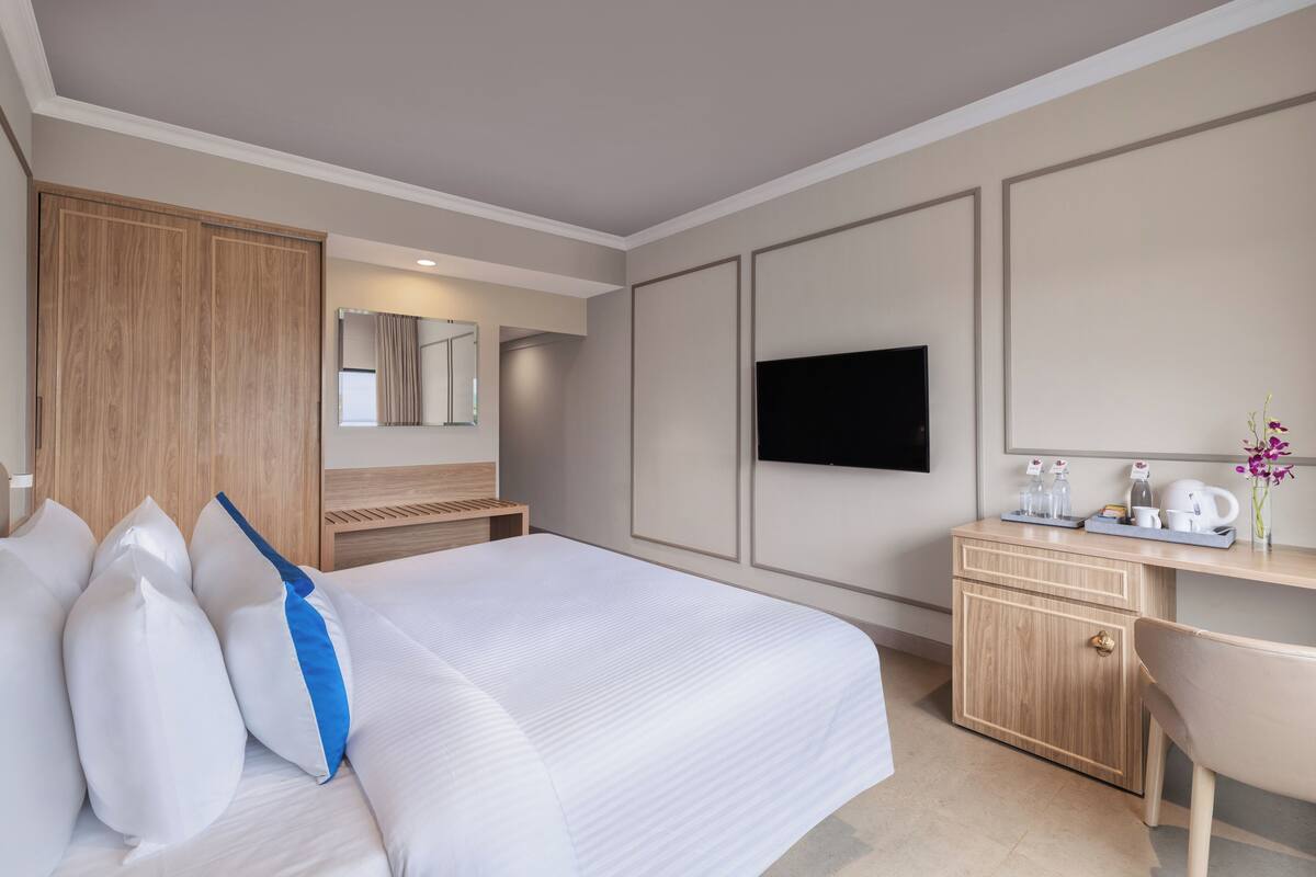 Quarto de hotel com uma cama grande, um guarda-roupa de madeira, uma mesa com cadeira, uma televisão e um espelho.