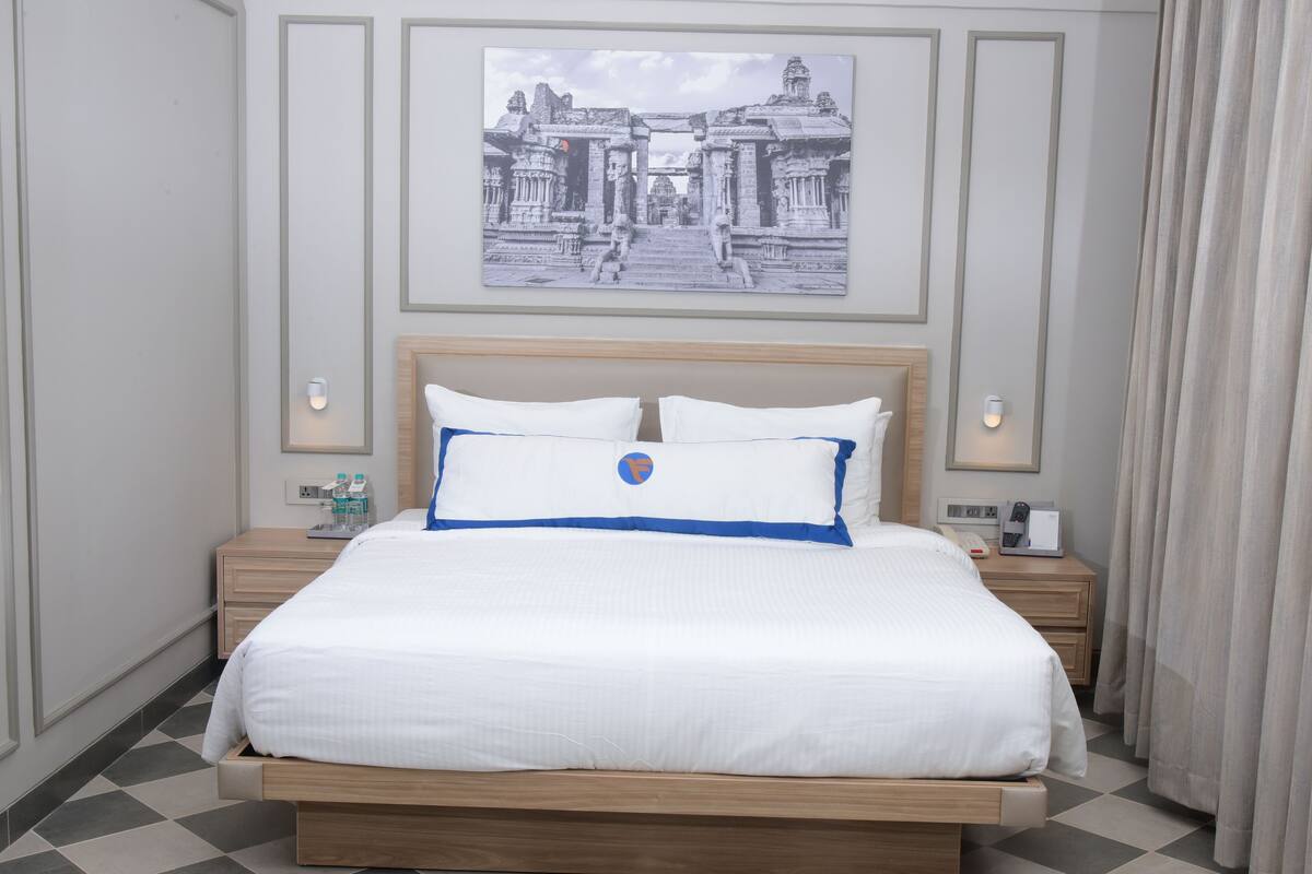 Cama bem arrumada com roupa de cama branca e detalhes azuis, uma moldura acima dela e uma mesa de cabeceira com abajur e telefone.