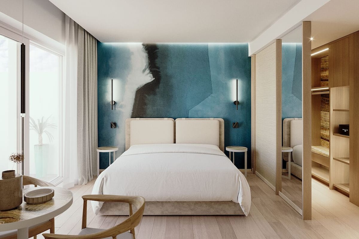 Quarto de hotel moderno com uma cama grande, mesas de cabeceira, área de estar com uma poltrona e vista para o exterior.