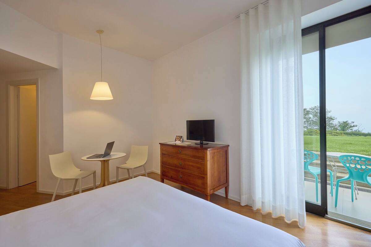 Quarto de hotel com uma cama, uma mesa com um laptop, uma televisão e vista para o exterior.