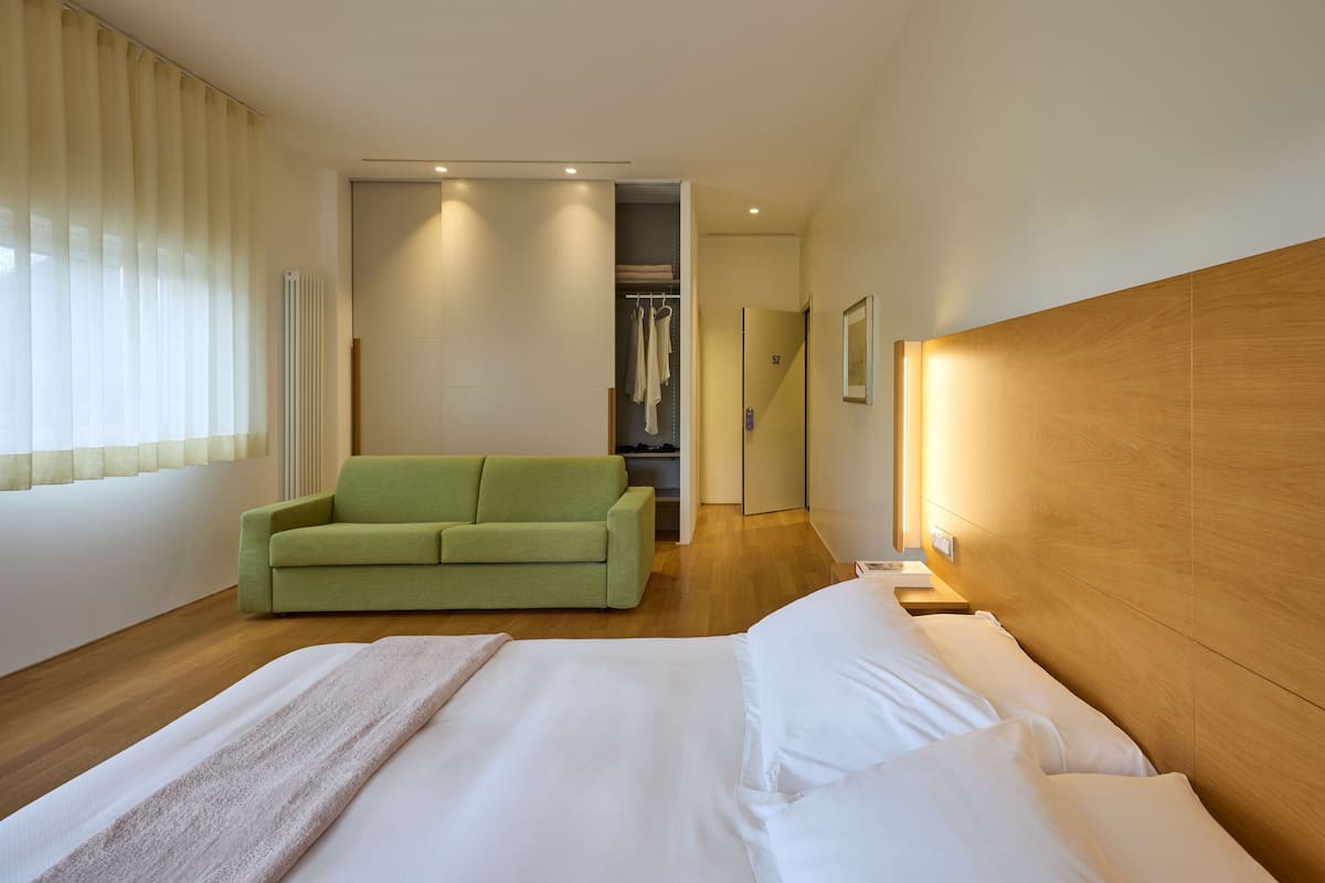 Quarto de hotel com cama, sofá verde, cabeceira de madeira e um closet espaçoso.