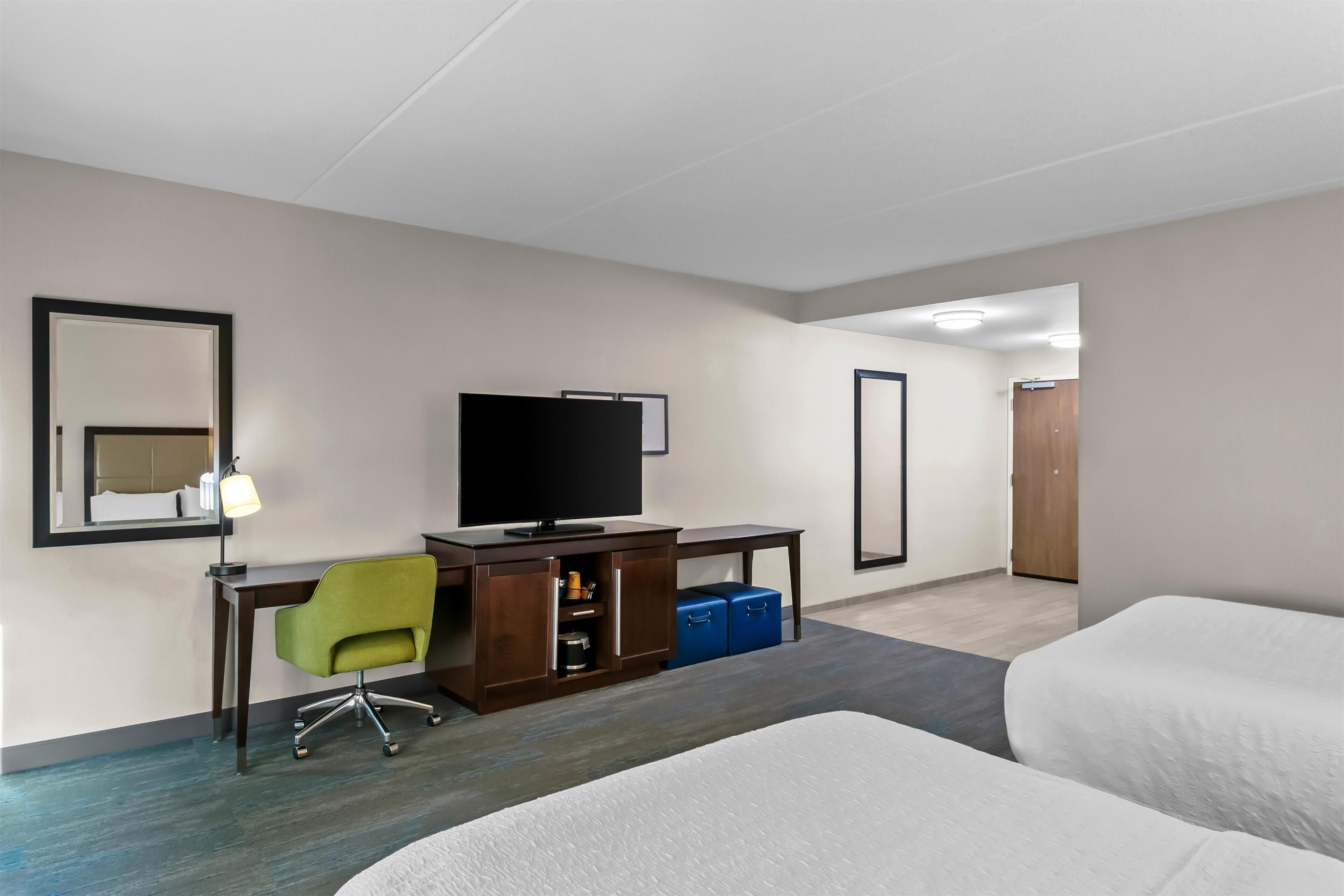 Quarto de hotel com cama, escrivaninha com cadeira, televisão e armário.