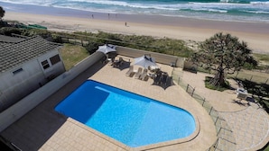 Piscina com espreguiçadeiras e guarda-sóis, vista para a praia e área cercada com uma árvore.