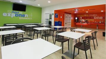 Uma cafeteria com mesas e cadeiras brancas, uma parede verde com um aviso, e uma parede laranja com um aviso.