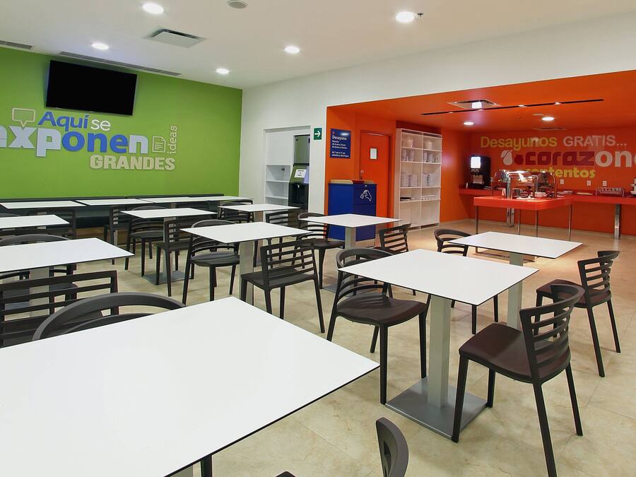 Uma cafeteria com mesas e cadeiras brancas, uma parede verde com um aviso, e uma parede laranja com um aviso.