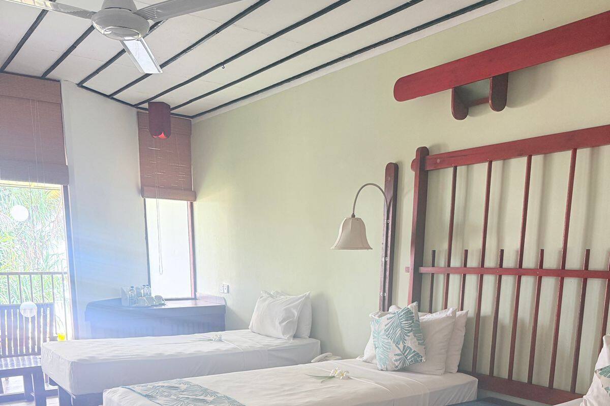 Quarto de hotel com duas camas, um ventilador de teto, janela com persianas e uma pequena mesa com abajur.