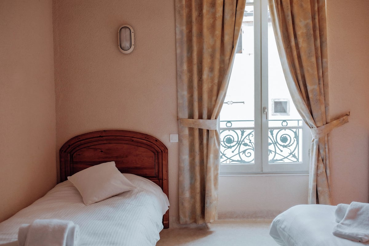 Quarto com cabeceira de madeira, janela com cortinas e varanda com grade de ferro forjado.