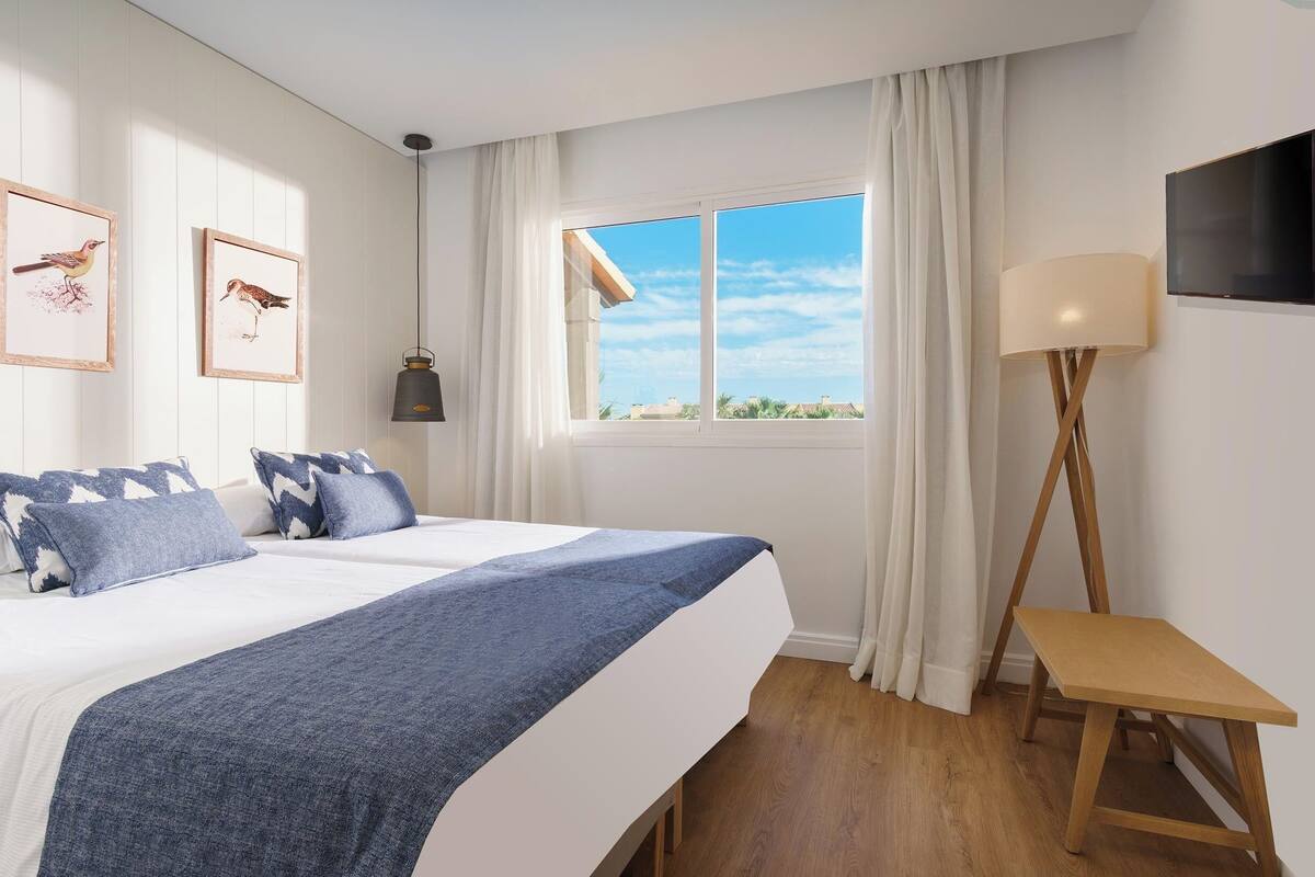 Quarto de hotel com uma cama grande, mesa de cabeceira de madeira, televisão na parede e janela com vista para edifícios.