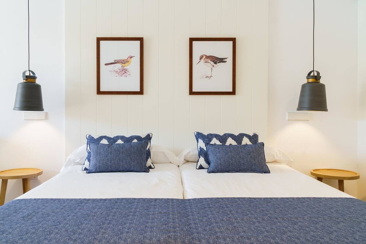 Uma cama de casal com duas almofadas, um cobertor azul e duas luminárias pendentes fixadas na parede.