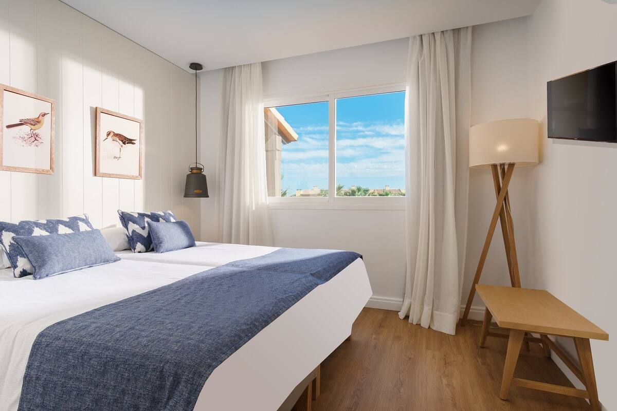 Quarto de hotel com uma cama grande, mesa de cabeceira de madeira, televisão na parede e janela com vista para edifícios.