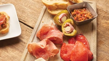 Tábua de corte de madeira com diversos alimentos, incluindo pão, tomates, picles e uma pequena tigela de salsa.