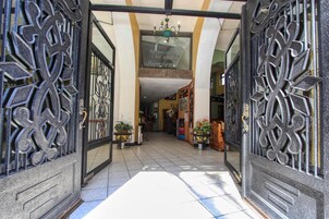Entrada de hotel com portões de ferro negro ornamentados, piso de azulejos e um candelabro.