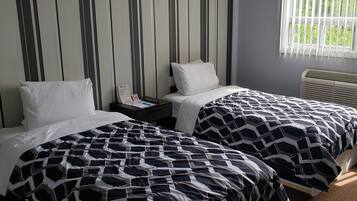 Quarto de hotel com duas camas, papel de parede listrado, mesa de cabeceira com telefone e papéis, e janela com persianas.