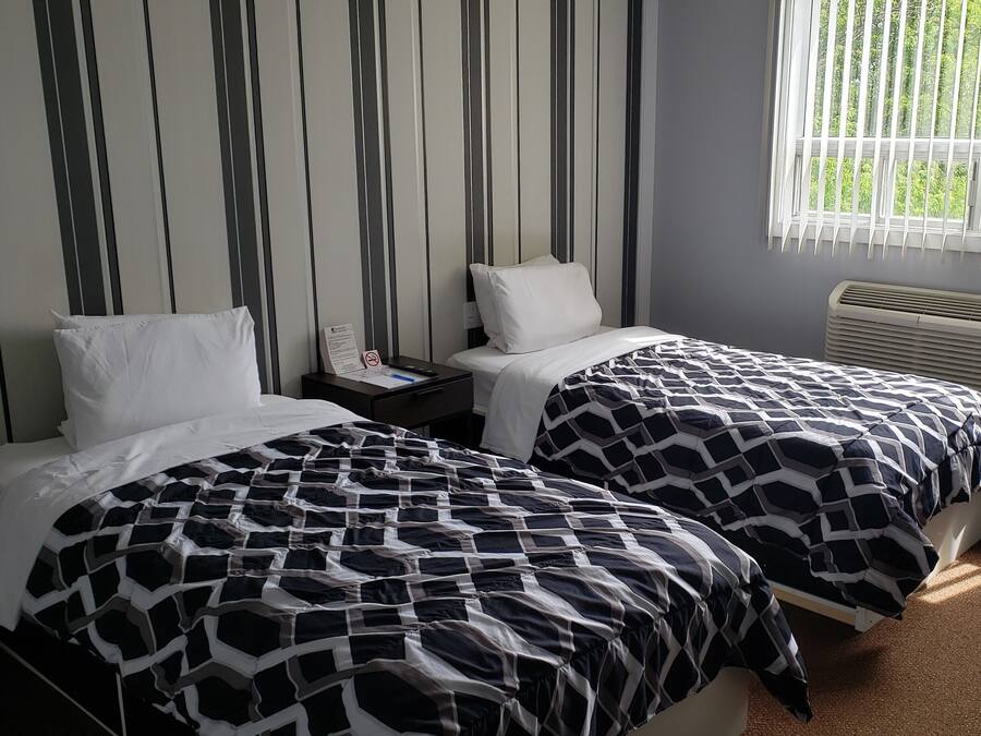 Quarto de hotel com duas camas, papel de parede listrado, mesa de cabeceira com telefone e papéis, e janela com persianas.