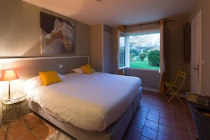 Quarto com uma cama grande, mesa de cabeceira com abajur, janela com vista para o verde e uma cadeira perto da janela.