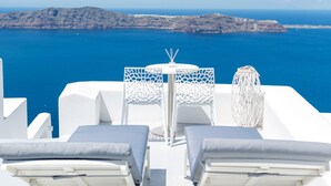 Terraço com cadeiras de estar brancas e uma mesa, com vista para um mar azul límpido e um litoral rochoso ao longe.