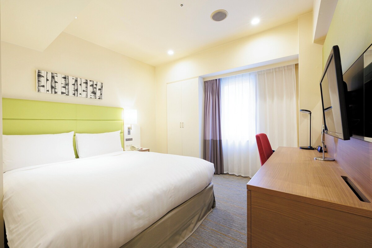 Quarto de hotel com uma cama grande, uma escrivaninha com televisão, uma cadeira, um guarda-roupa e uma janela com cortinas.