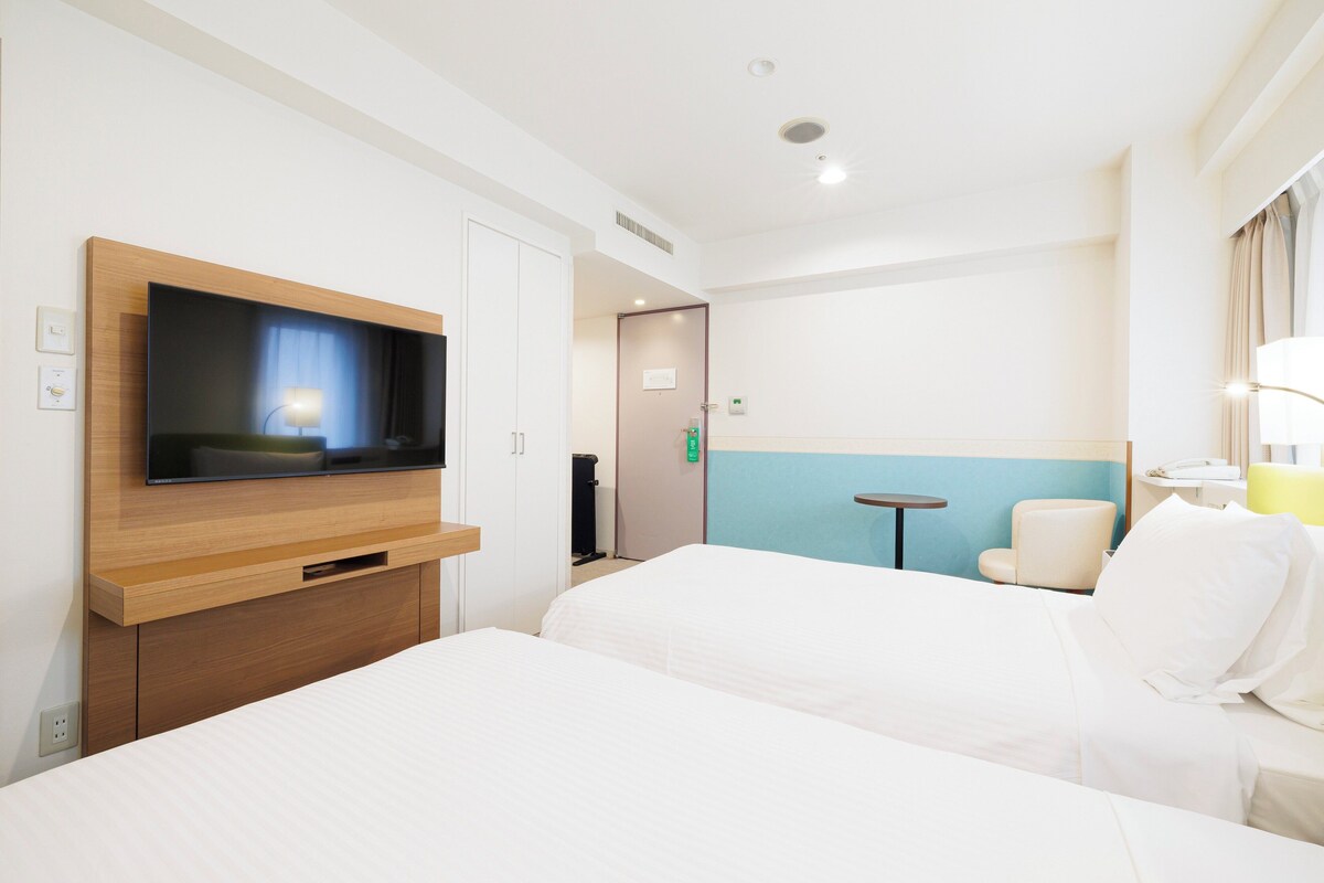 Quarto de hotel com duas camas, uma televisão fixada na parede, uma mesinha e uma cadeira.