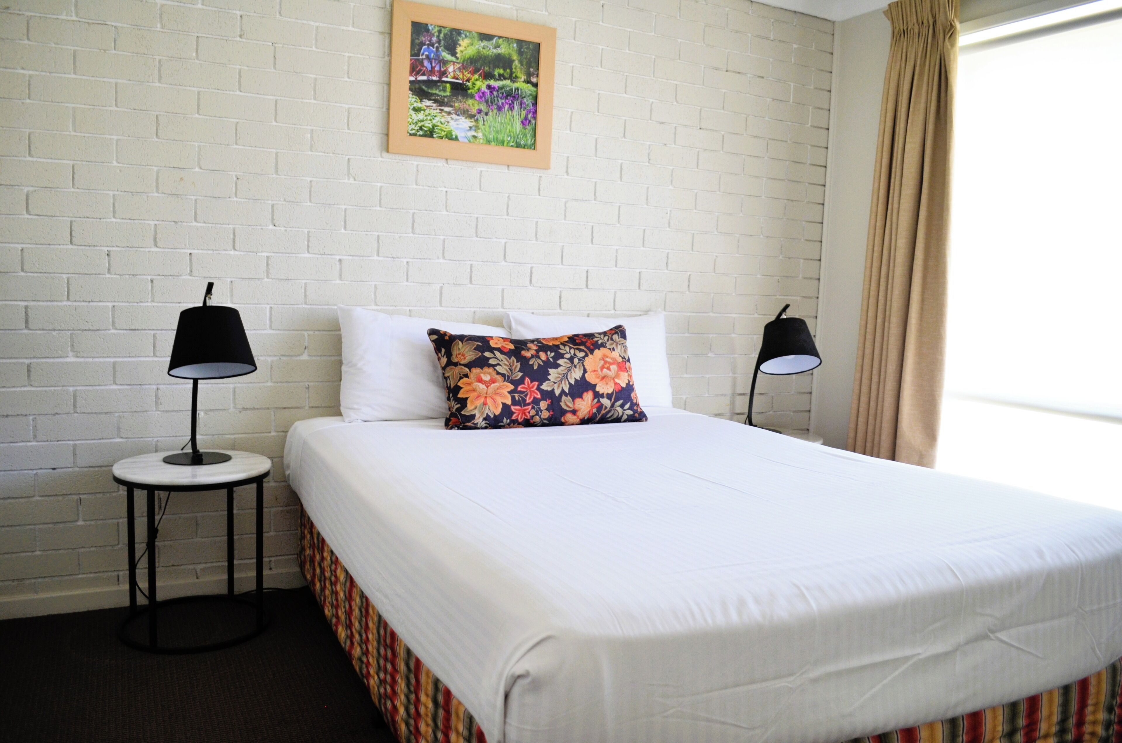 Cama bem arrumada com roupa de cama branca e almofada floral, ladeada por duas luminárias pretas sobre mesas redondas, em frente a uma parede de tijolos brancos com um quadro emoldurado acima.